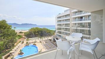 Varanda com vista para piscina e praia, cadeiras brancas e guarda-corpo de vidro.