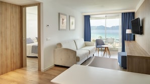 Quarto de hotel moderno com piso de madeira, sofá branco, mesa de jantar e TV fixada na parede.