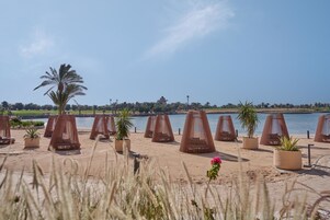 Uma praia com espreguiçadeiras e plantas em vasos, uma palmeira e um corpo d'água ao fundo.