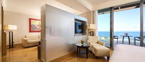 Quarto de hotel moderno com uma cama grande, um sofá, uma poltrona, uma televisão e varanda com vista para o mar.