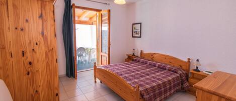 Quarto com cama de madeira, guarda-roupa de madeira, mesa de cabeceira pequena com luminária e vista para uma varanda através de uma porta aberta.