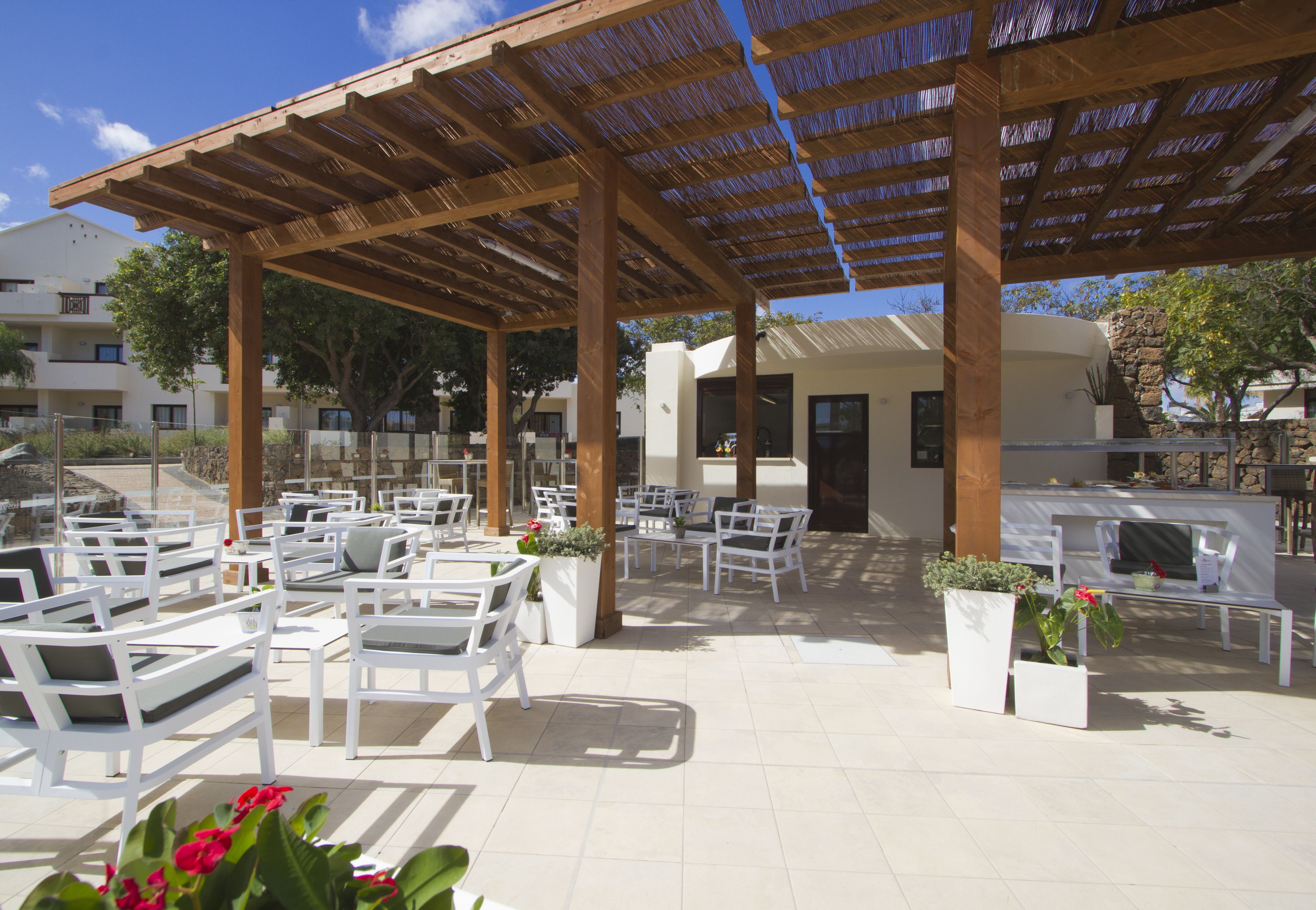 Um pátio externo com cadeiras e mesas brancas, uma pérgola de madeira e plantas em vasos.