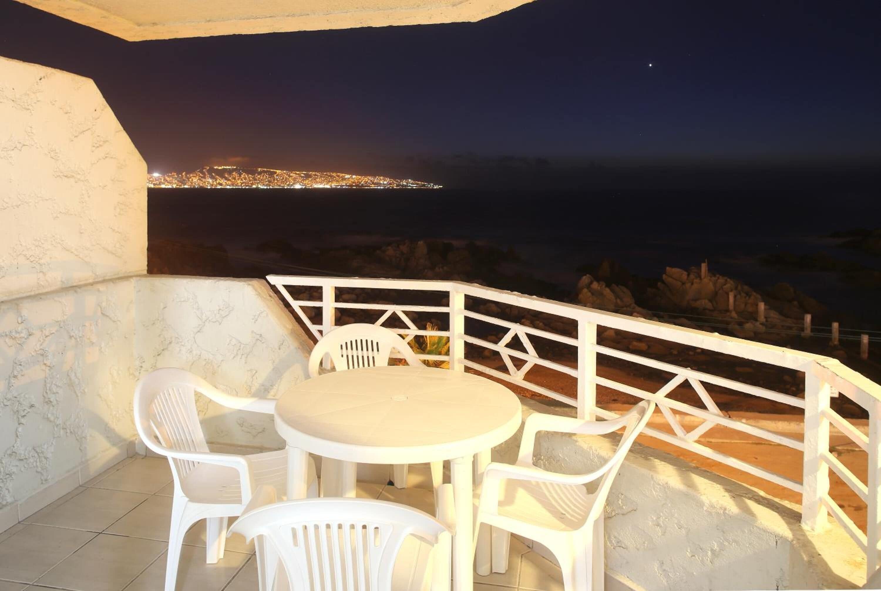 Uma varanda com mesa e cadeiras brancas, com vista para a cidade iluminada à noite.