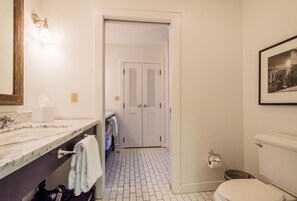 Um banheiro com pia de mármore, piso de azulejos brancos, vaso sanitário branco e um quadro pendurado na parede.