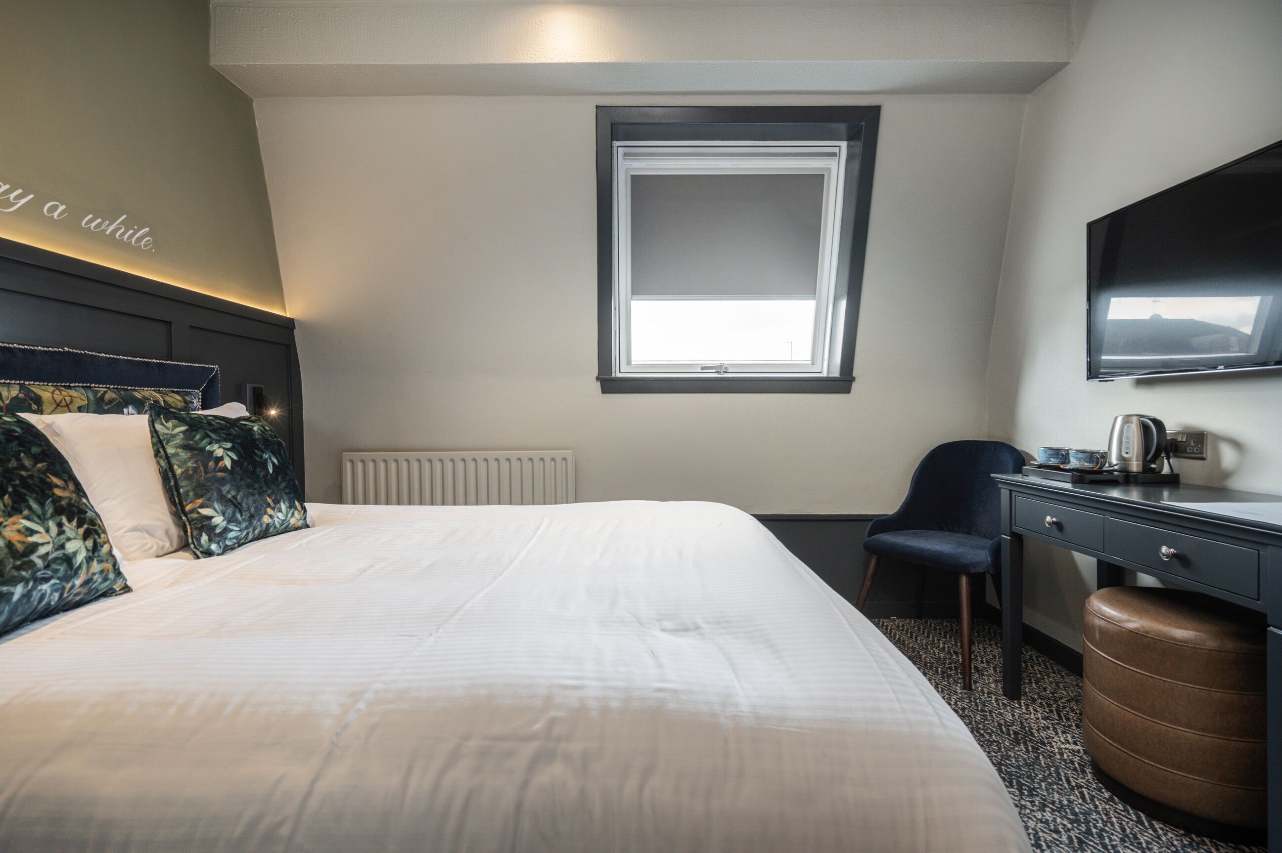 Quarto de hotel com cama, escrivaninha, cadeira, janela e televisão.
