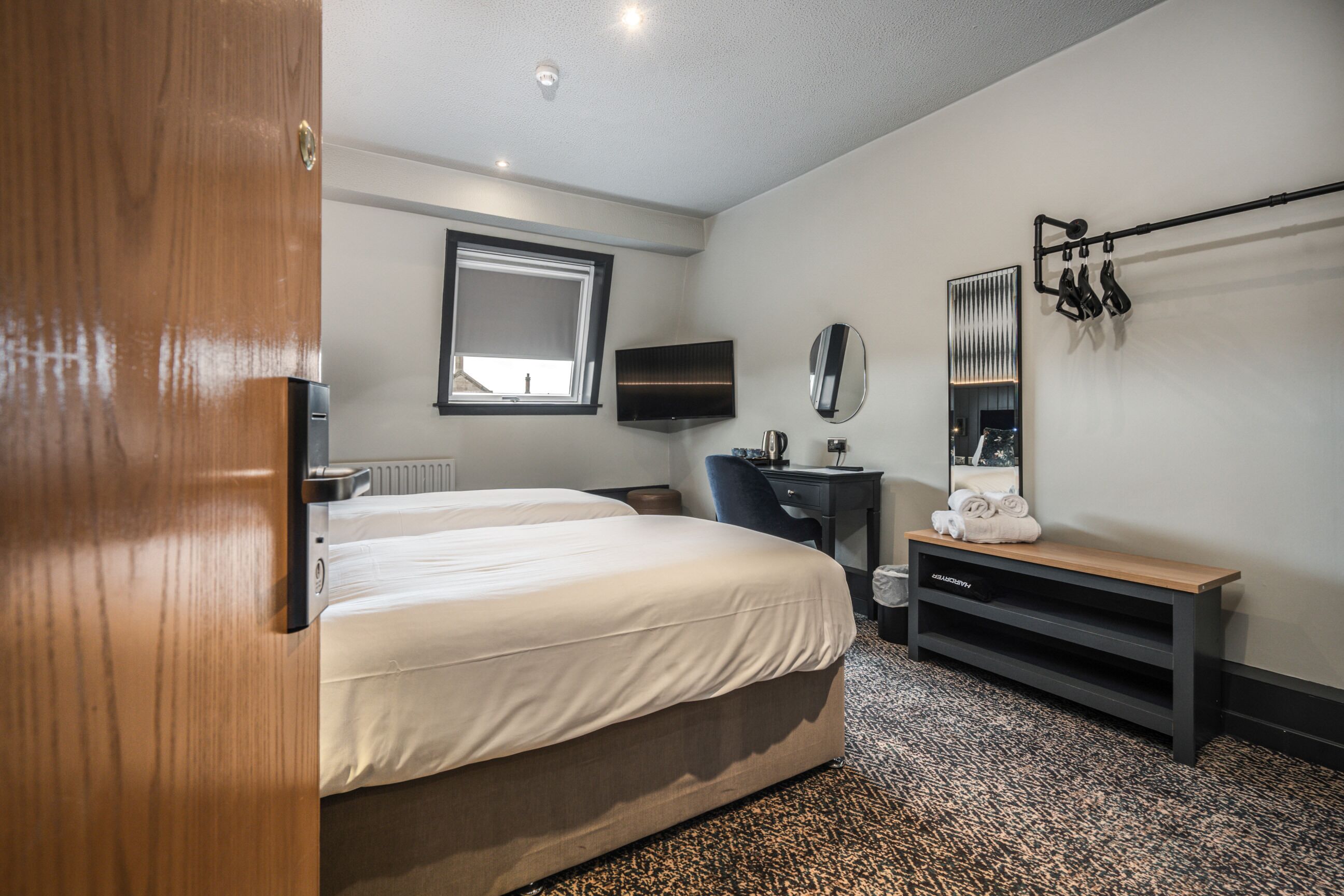 Quarto de hotel com uma cama grande, uma escrivaninha, uma cadeira, uma televisão, um espelho e um sapateiro.