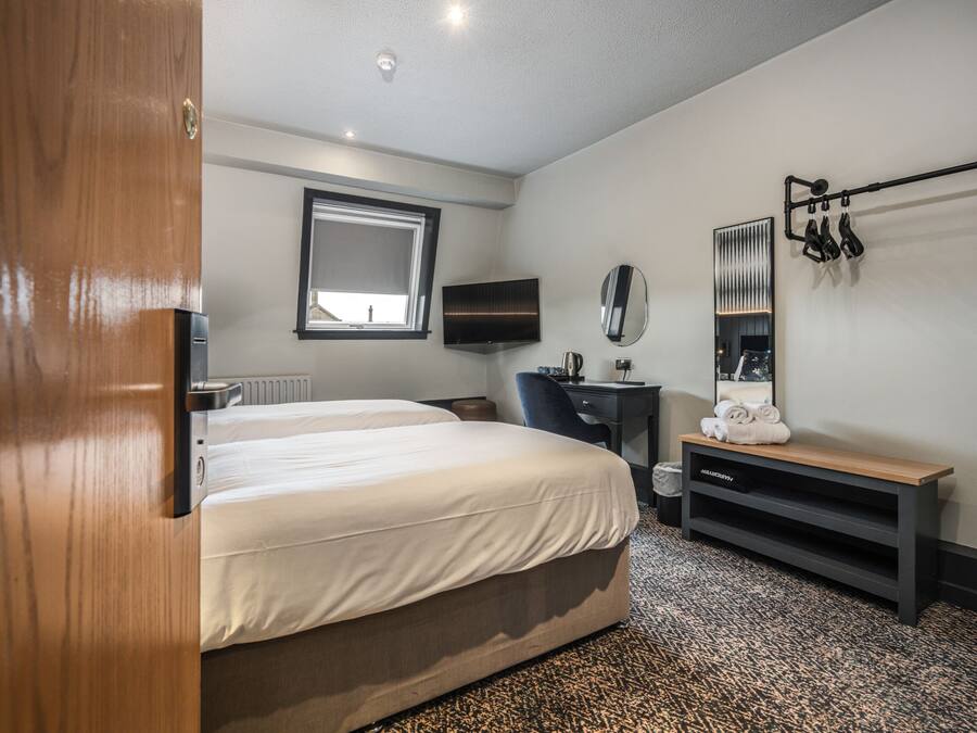 Quarto de hotel com uma cama grande, uma escrivaninha, uma cadeira, uma televisão, um espelho e um sapateiro.