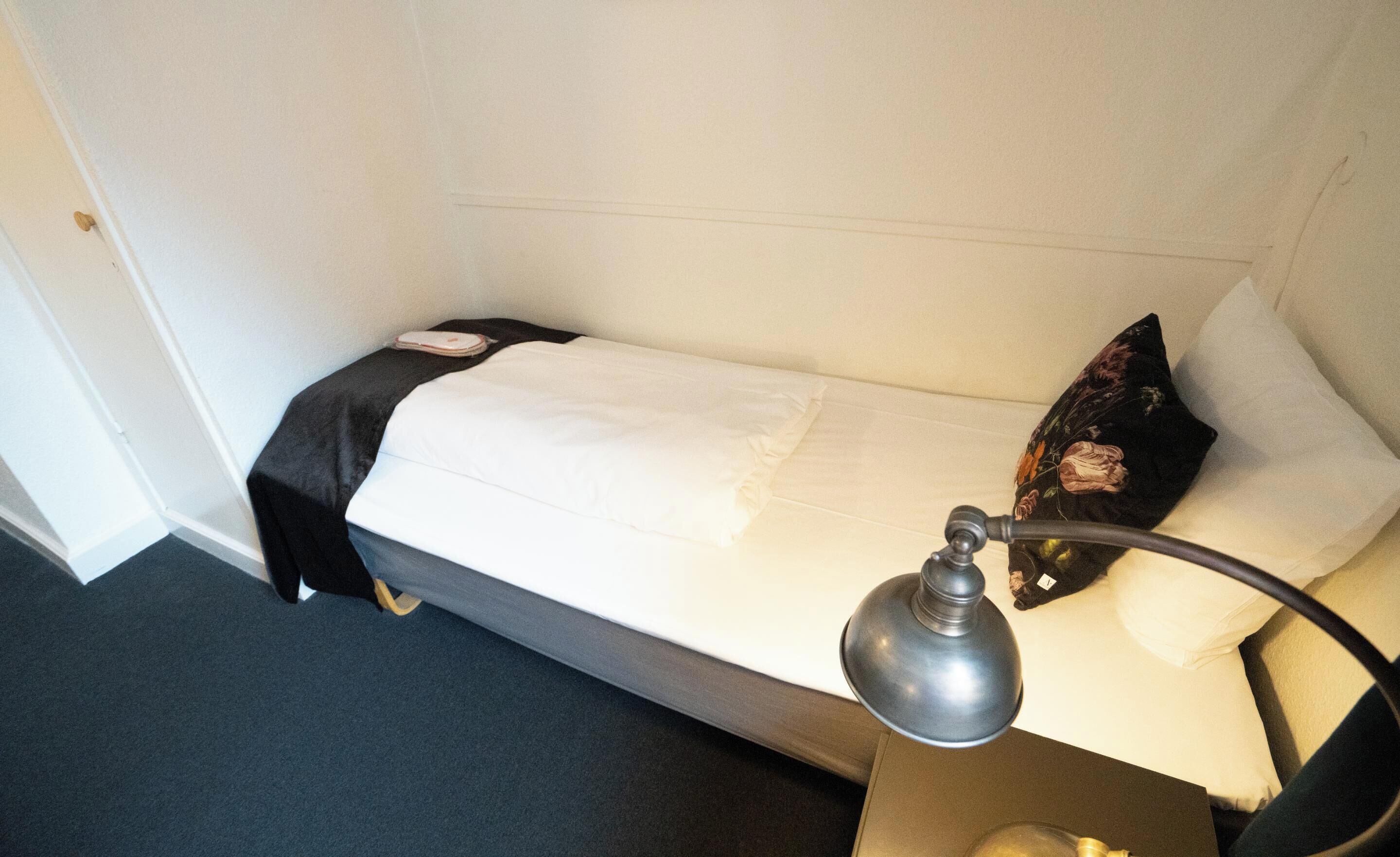 Quarto pequeno com uma cama de solteiro, mesa de cabeceira, abajur e travesseiro.