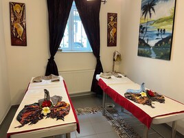 Um quarto com duas mesas de massagem, cada uma com toalhas e uma flor. Há cortinas, uma janela e obras de arte nas paredes.