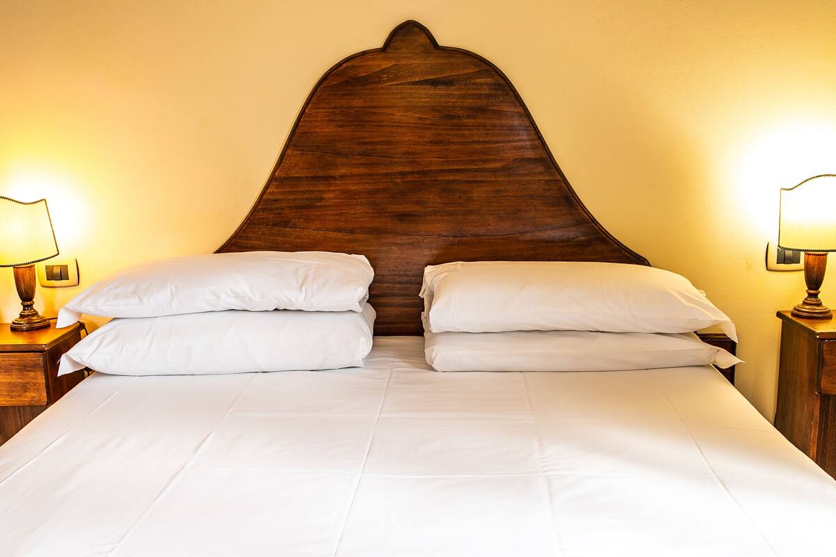 Uma cama de casal com cabeceira de madeira, duas mesas de cabeceira com abajures e roupa de cama branca.