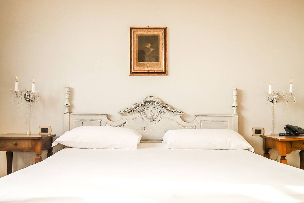 Cama bem arrumada com lençóis brancos, uma cabeceira clássica e ornamentada, e duas mesas de cabeceira com luminárias e um relógio.