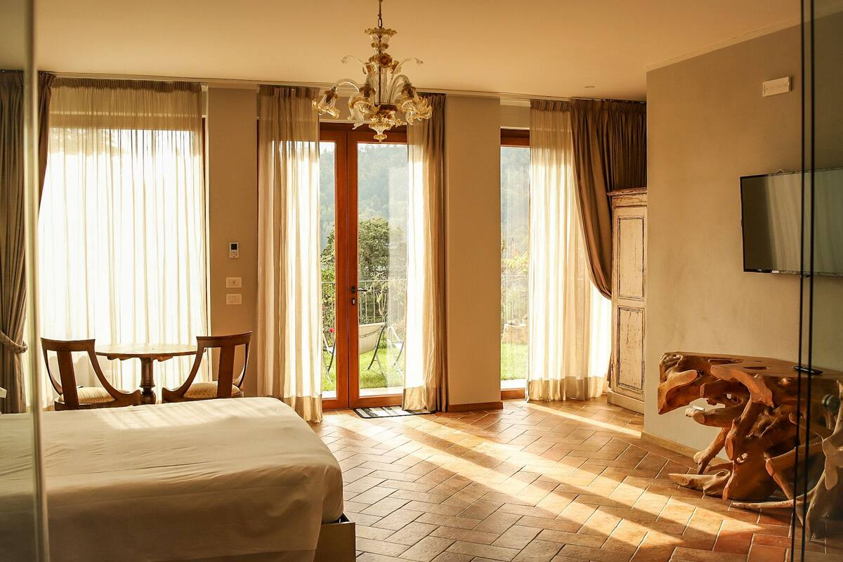 Quarto de hotel com cama, mesa de jantar com cadeiras, lustre, televisão e janelas amplas com cortinas.