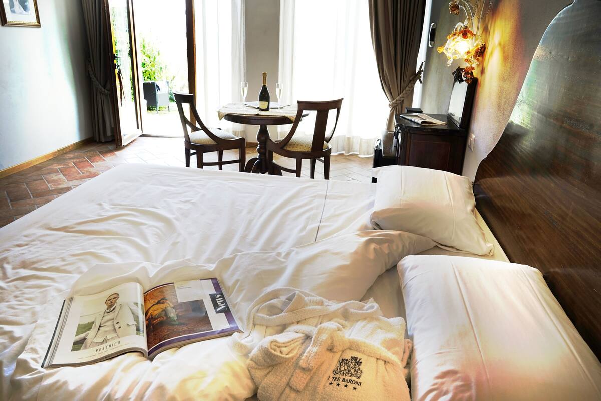 Quarto de hotel com uma cama grande, uma mesa de cabeceira com garrafa e copos, uma cadeira e vista para o exterior.