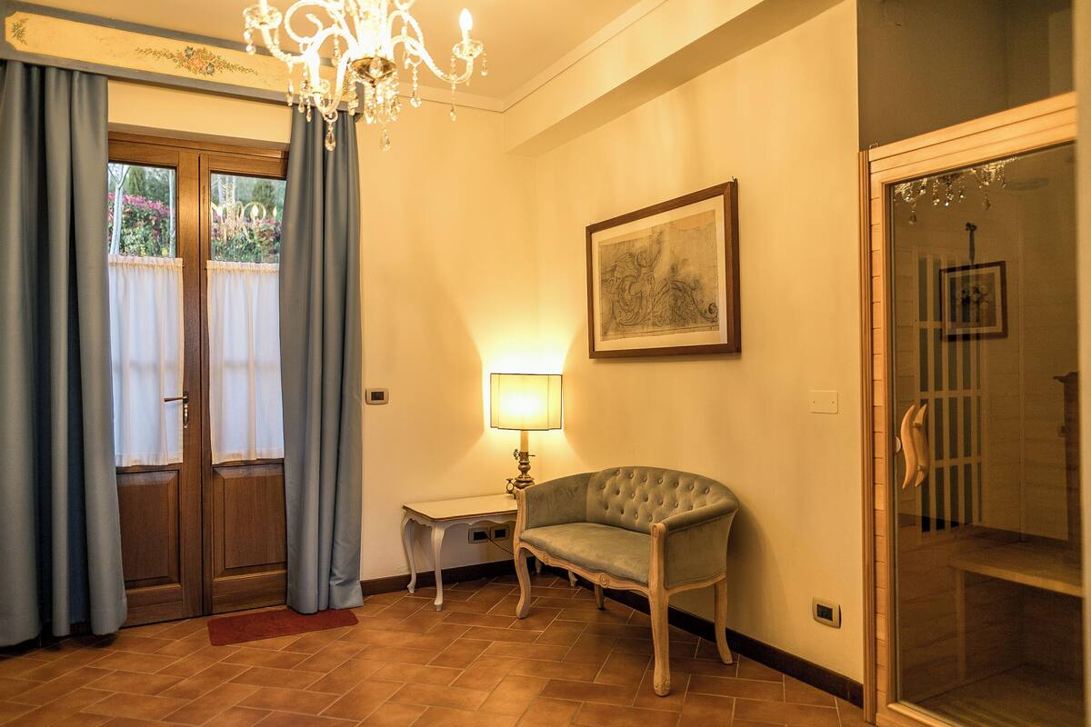 Quarto com porta de madeira, poltrona estofada, mesa pequena com abajur, quadro emoldurado na parede e porta de vidro para área do chuveiro.