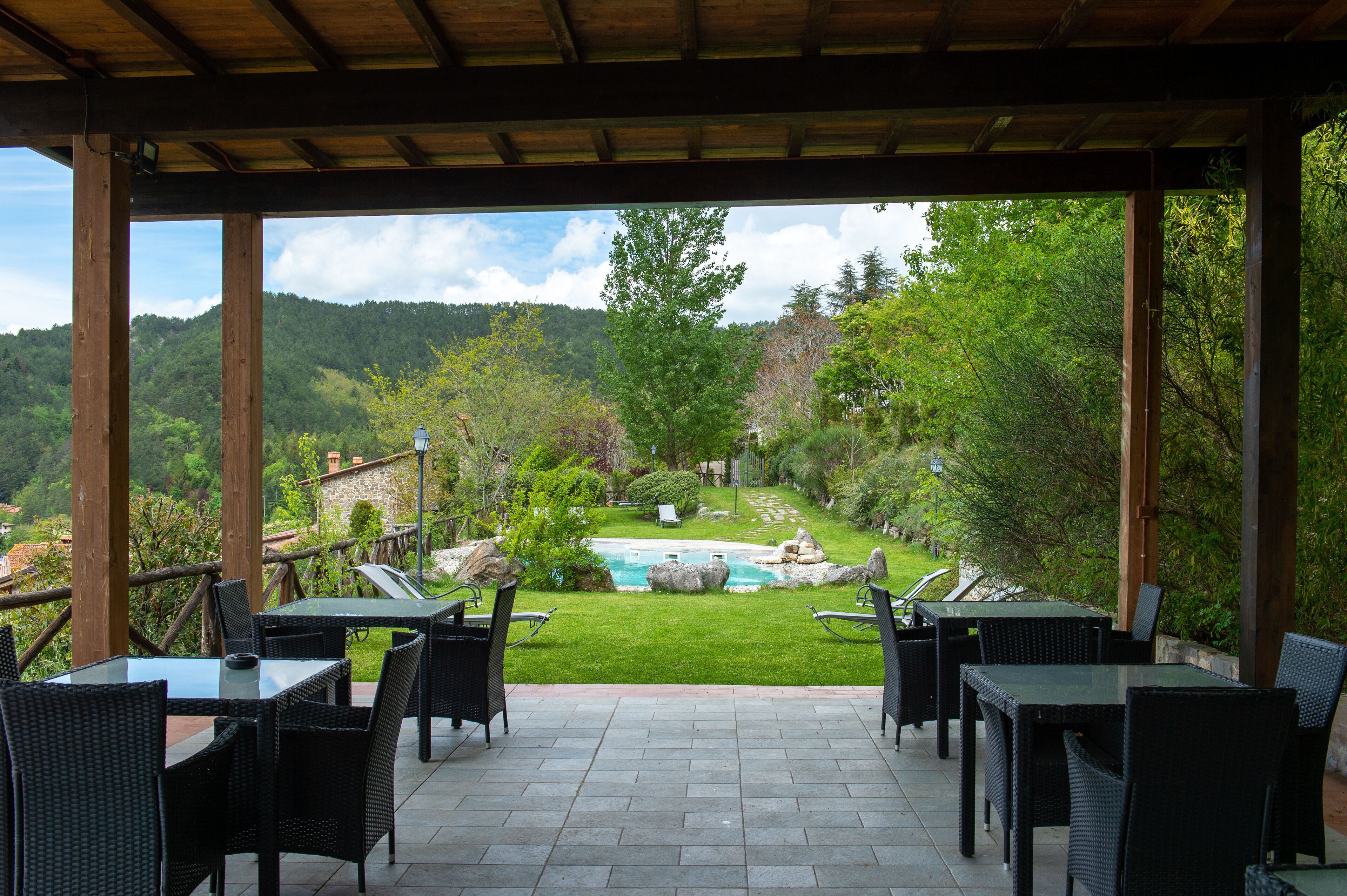 Um pátio externo com mesas e cadeiras, uma piscina e vista para um jardim exuberante e colinas ao fundo.