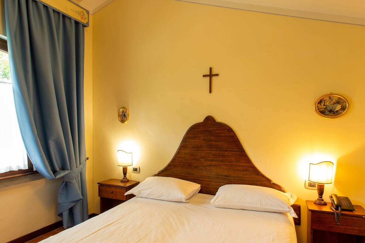 Quarto de hotel com uma cama, duas luminárias de cabeceira, um painel de cabeceira de madeira, uma cruz na parede e uma janela com cortinas.