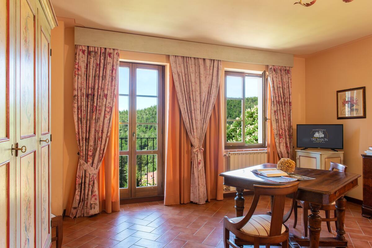 Uma área de refeições com uma mesa e cadeiras de madeira, uma televisão em um suporte e vista para vegetação pelas janelas.