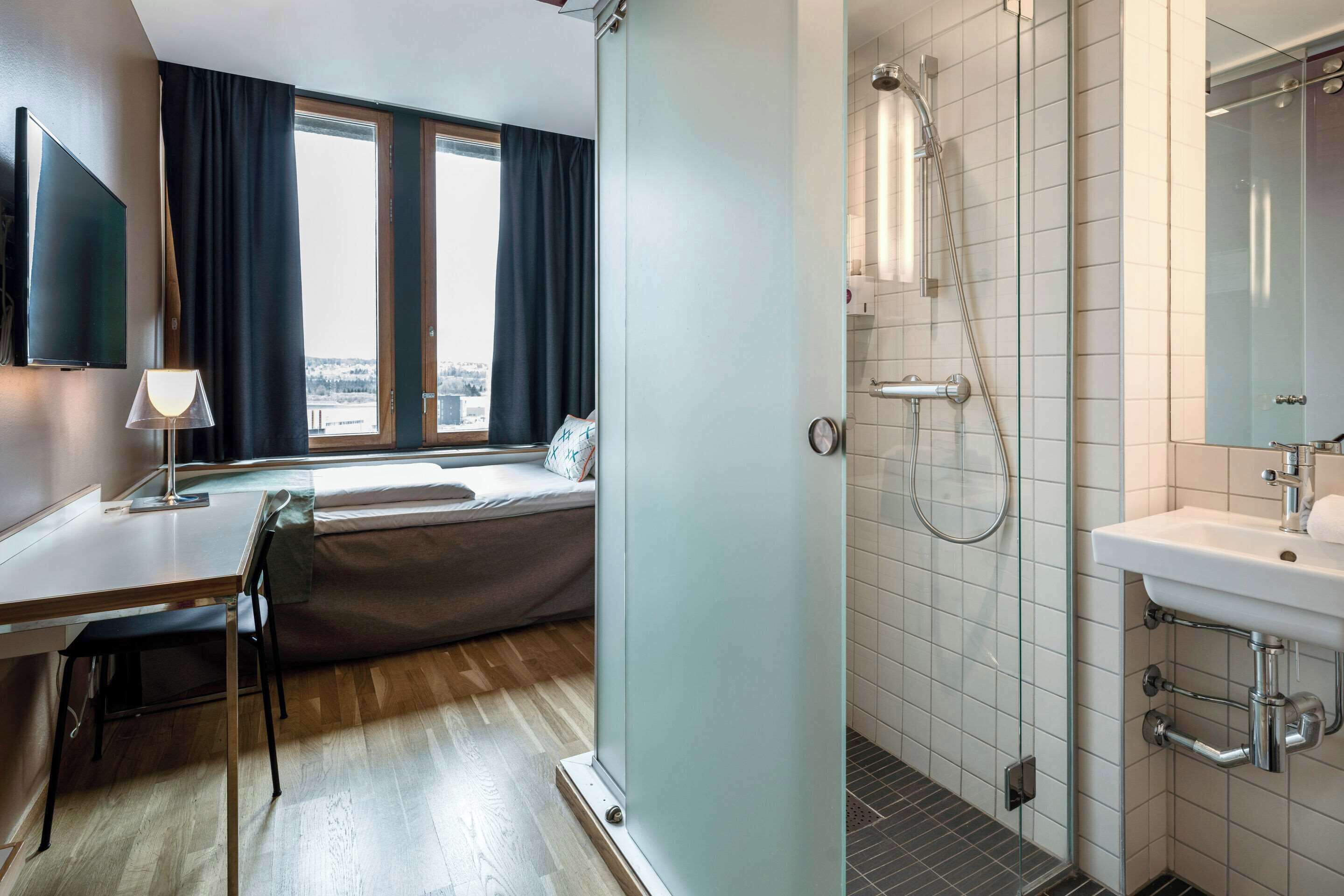 Quarto de hotel moderno com cama, escrivaninha, poltrona, televisão, janela com cortinas e chuveiro.