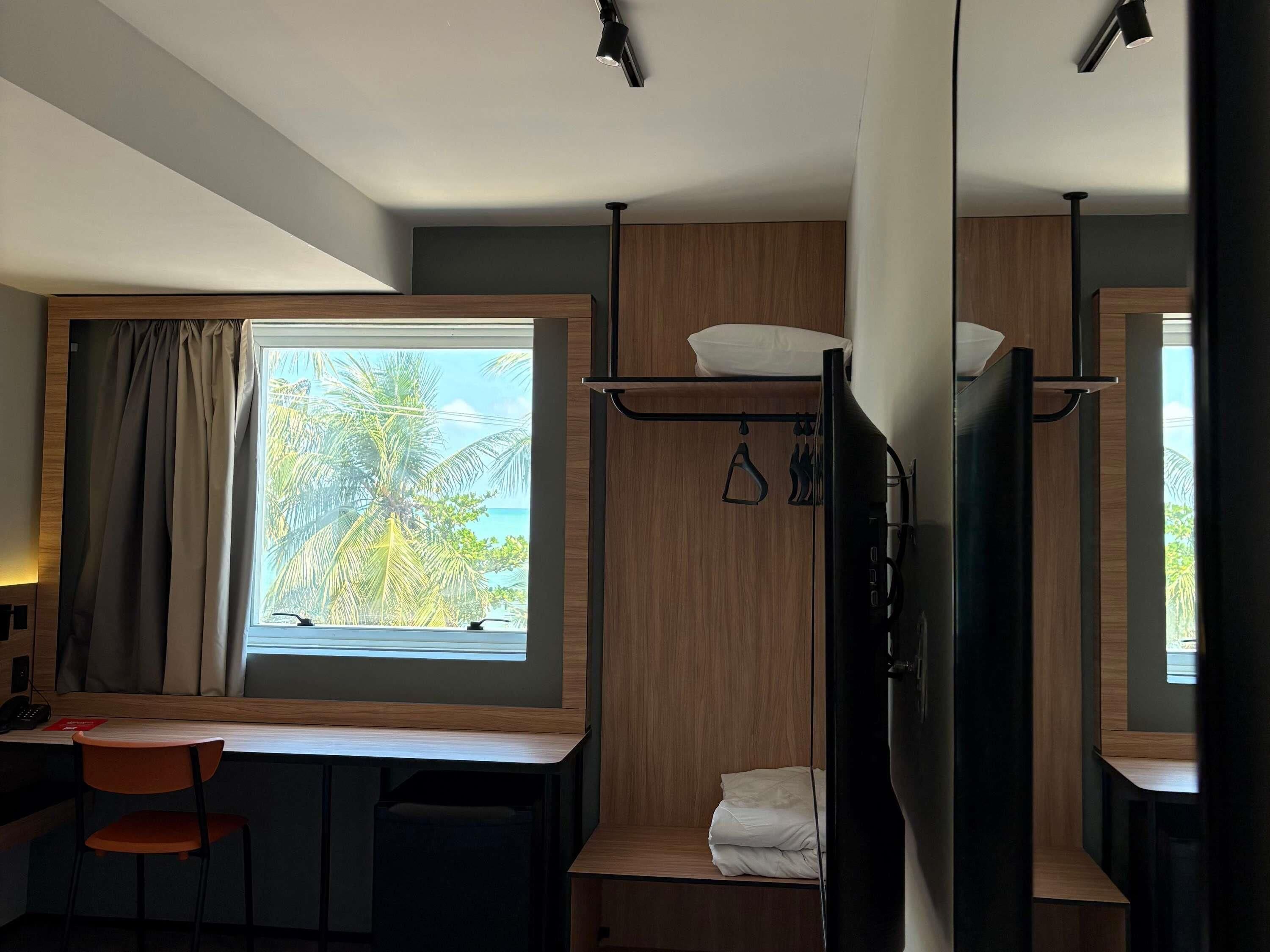 Quarto moderno com escrivaninha, cadeira e cama, com vista para palmeiras e para o mar.