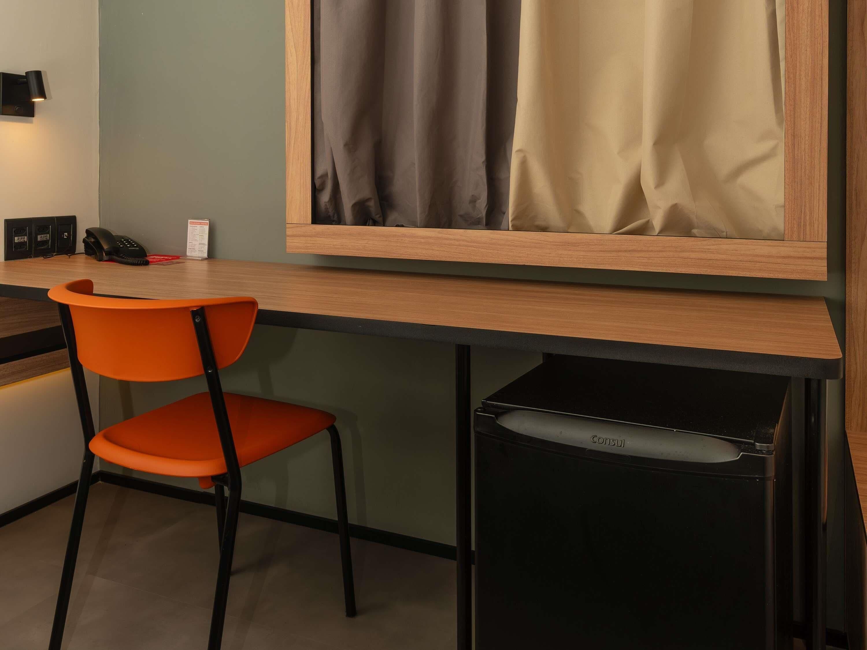 Quarto de hotel compacto com escrivaninha, cadeira, telefone, pequena geladeira preta e uma janela grande com cortinas.