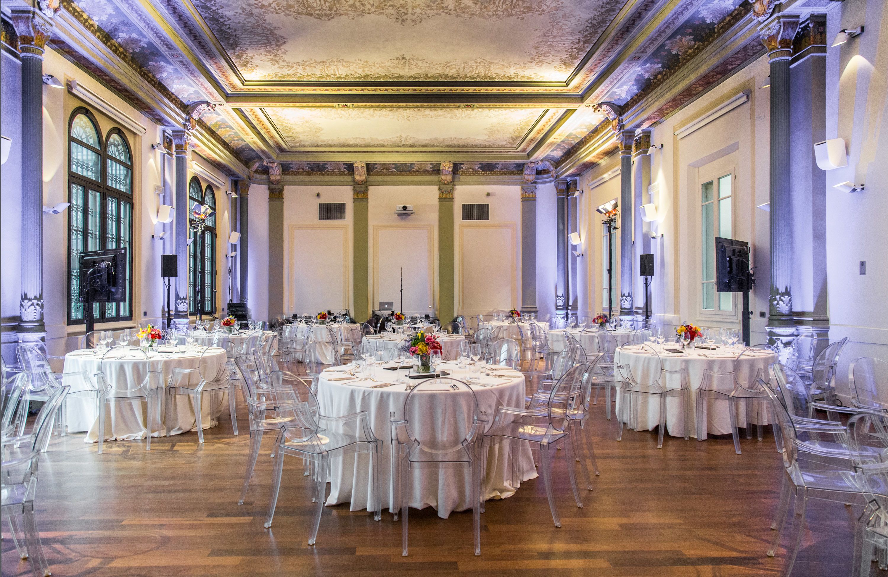 Uma sala de banquetes com mesas redondas, cadeiras transparentes e lustres.