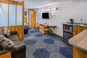 Quarto de hotel compacto com cozinha compacta, uma cama com cortina colorida, uma escrivaninha com duas cadeiras e um espelho grande.