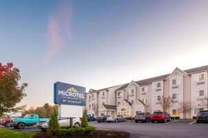 Um Microtel Inn & Suites by Wyndham com um estacionamento cheio de carros, um edifício com várias janelas e uma bandeira, sob um céu claro.