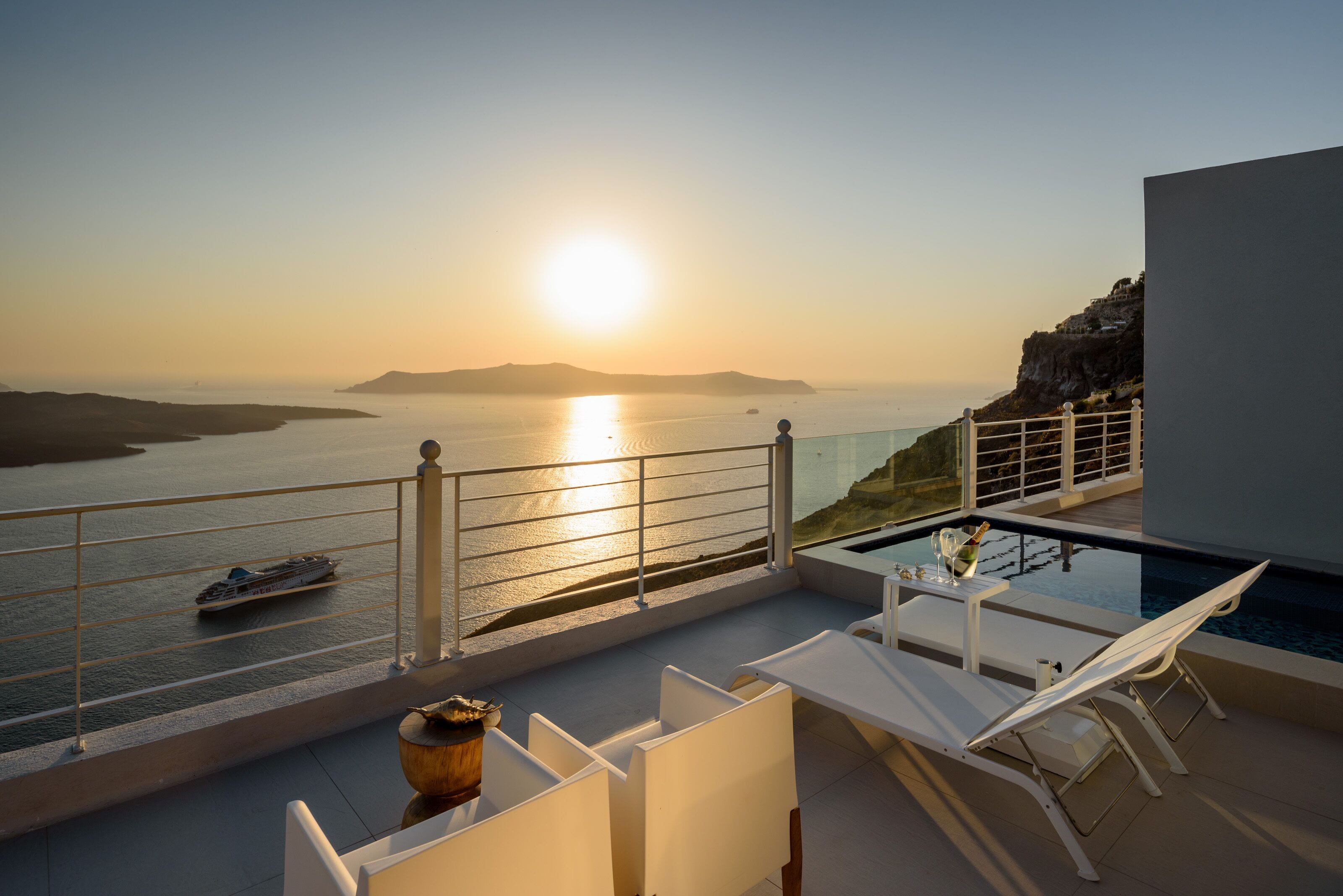 Varanda com vista para o mar e para o pôr do sol, piscina e cadeiras de sol brancas.