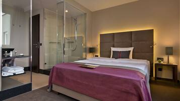 Quarto de hotel moderno com uma cama grande, box de chuveiro de vidro, toucador com artigos de higiene e mesa de cabeceira com abajur.
