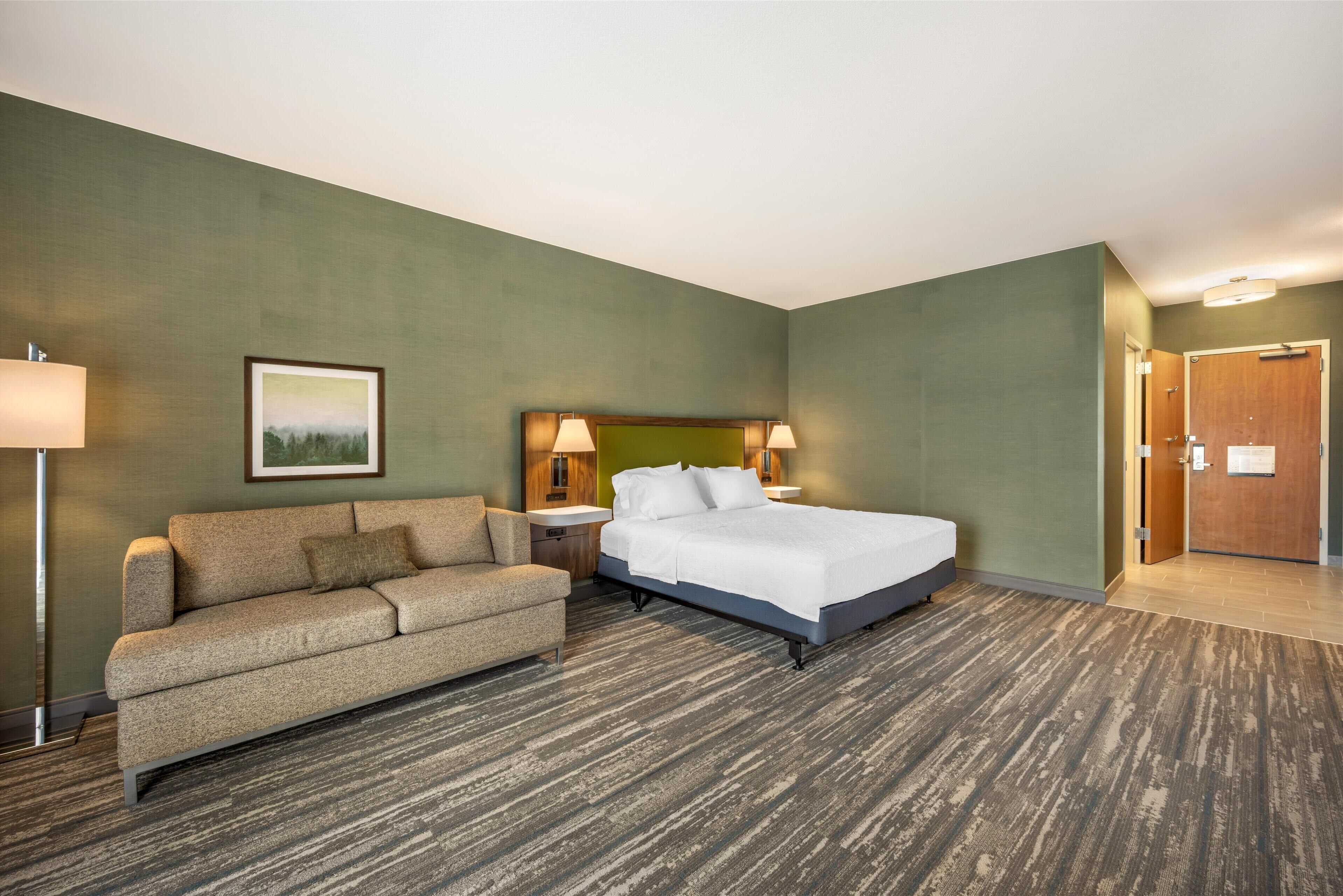 Quarto de hotel com cama, sofá, mesa de cabeceira e luminária fixada na parede.