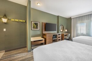 Quarto de hotel com duas camas, uma escrivaninha com cadeira, televisão, espelho e janela com cortinas.