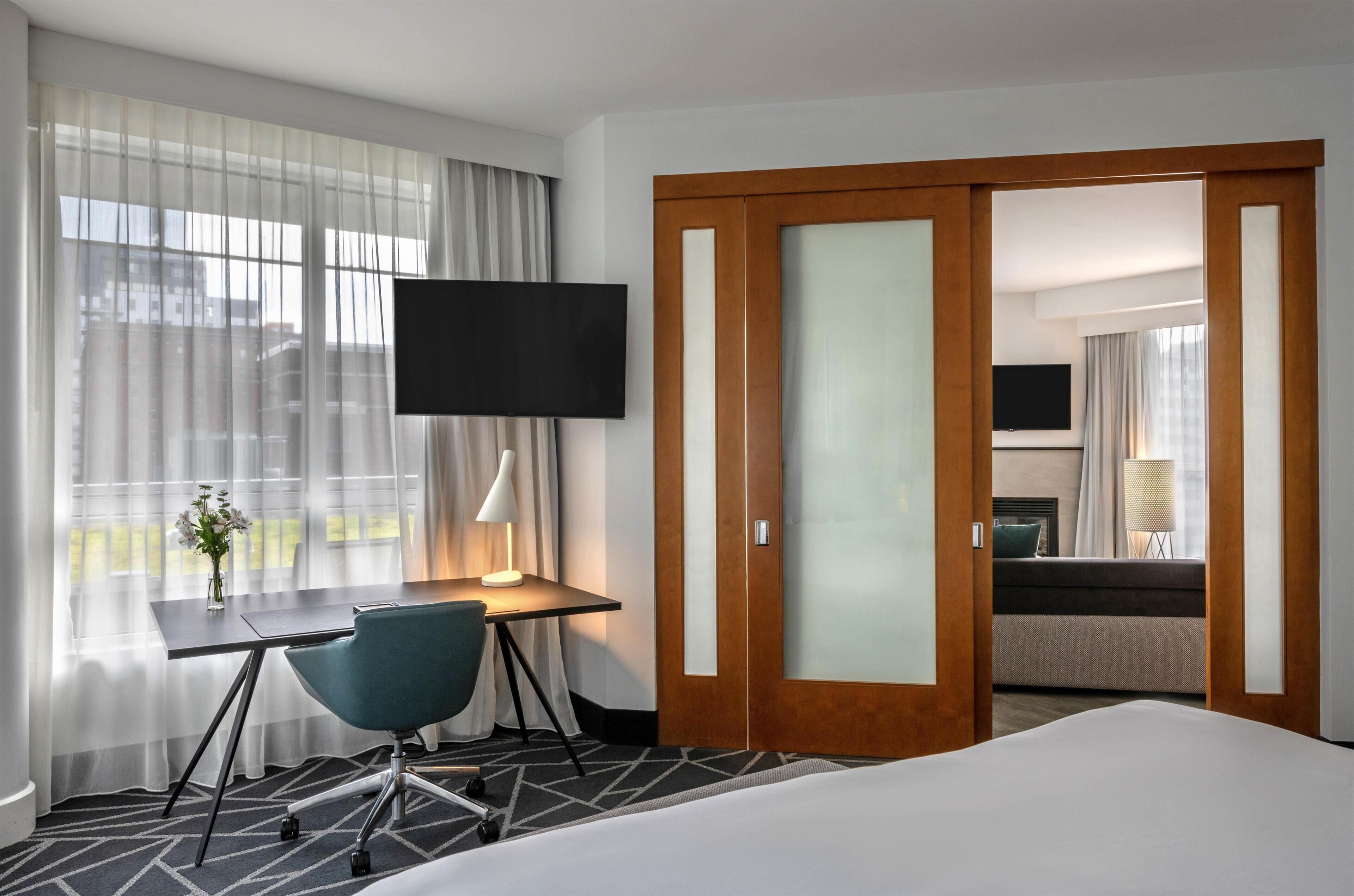 Quarto de hotel com cama, escrivaninha, cadeira, televisão e vista para o exterior pela janela.