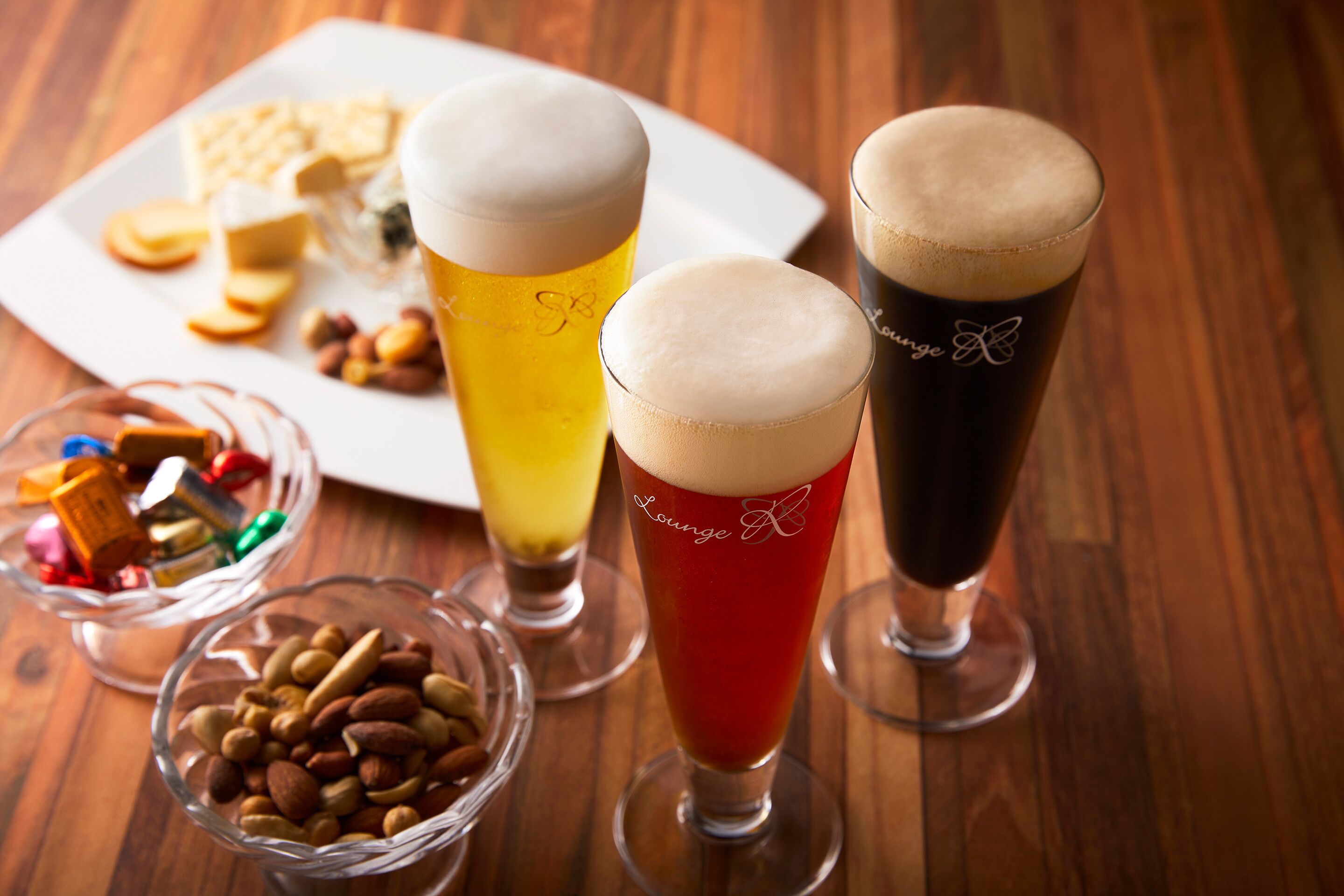 Três copos de cerveja sobre uma mesa de madeira, com um prato de petiscos e uma tigela de nozes.
