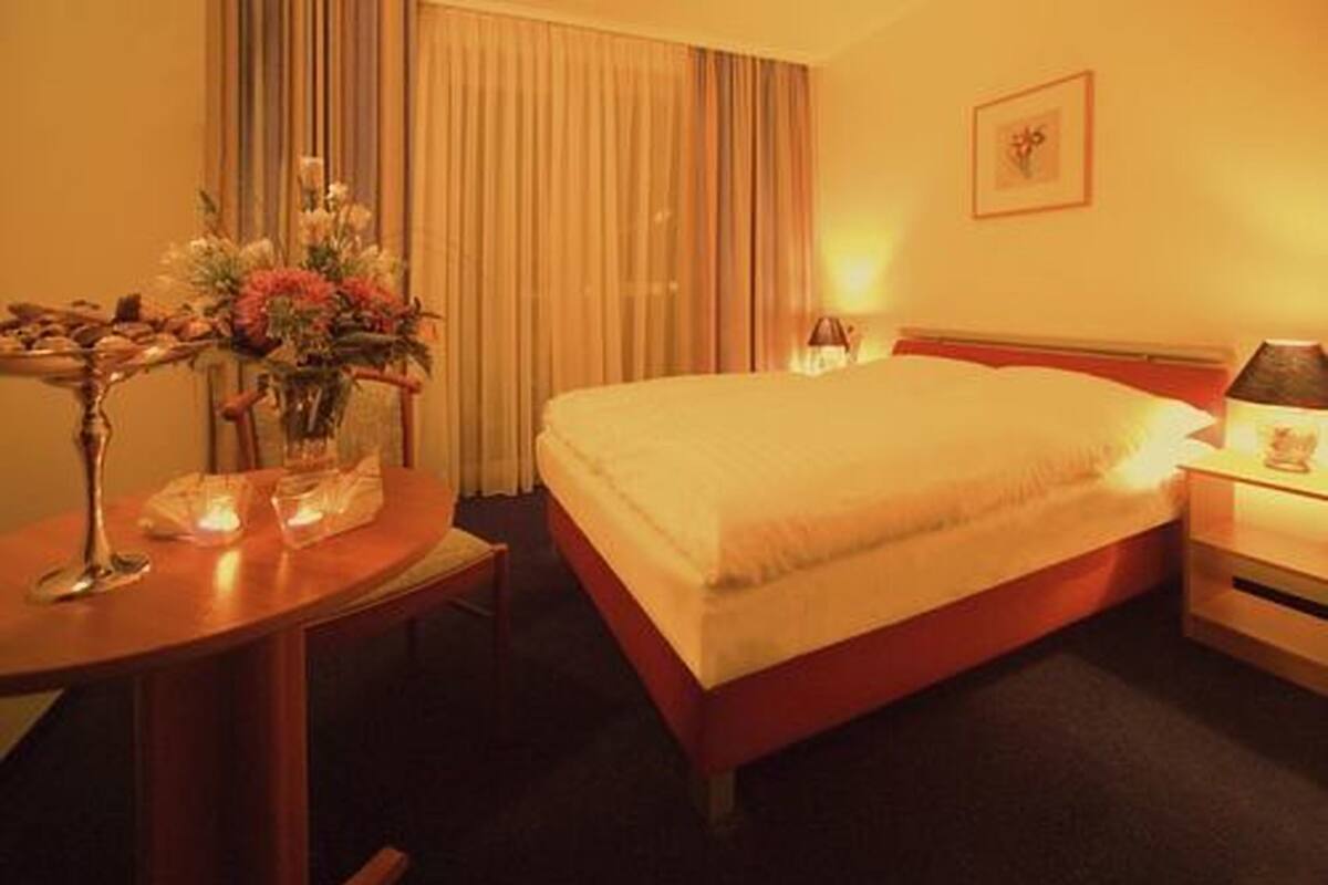 Quarto de hotel com cama, mesa de cabeceira, luminária, cadeira e mesa com um vaso de flores.