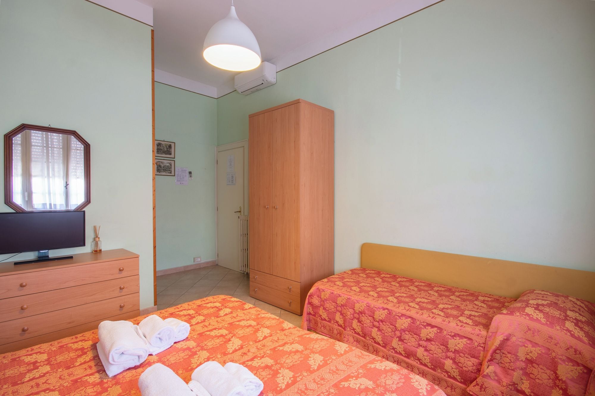 Quarto com duas camas, um guarda-roupa de madeira, uma televisão em uma cômoda e uma luminária no teto.