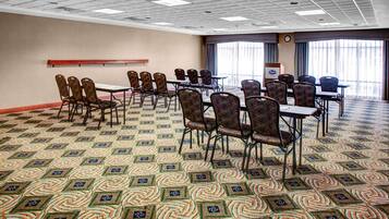 Uma sala de conferências com carpete estampado, fileiras de cadeiras, mesas, um relógio e uma janela com cortinas.