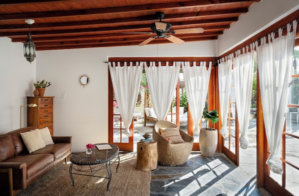 Uma sala de estar acolhedora com um sofá de couro marrom, uma mesa de centro redonda e uma cadeira de vime. Há um ventilador de teto, um armário de madeira e uma planta em vaso.