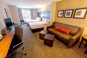 Quarto de hotel com duas camas, um sofá, uma escrivaninha com computador e uma televisão.