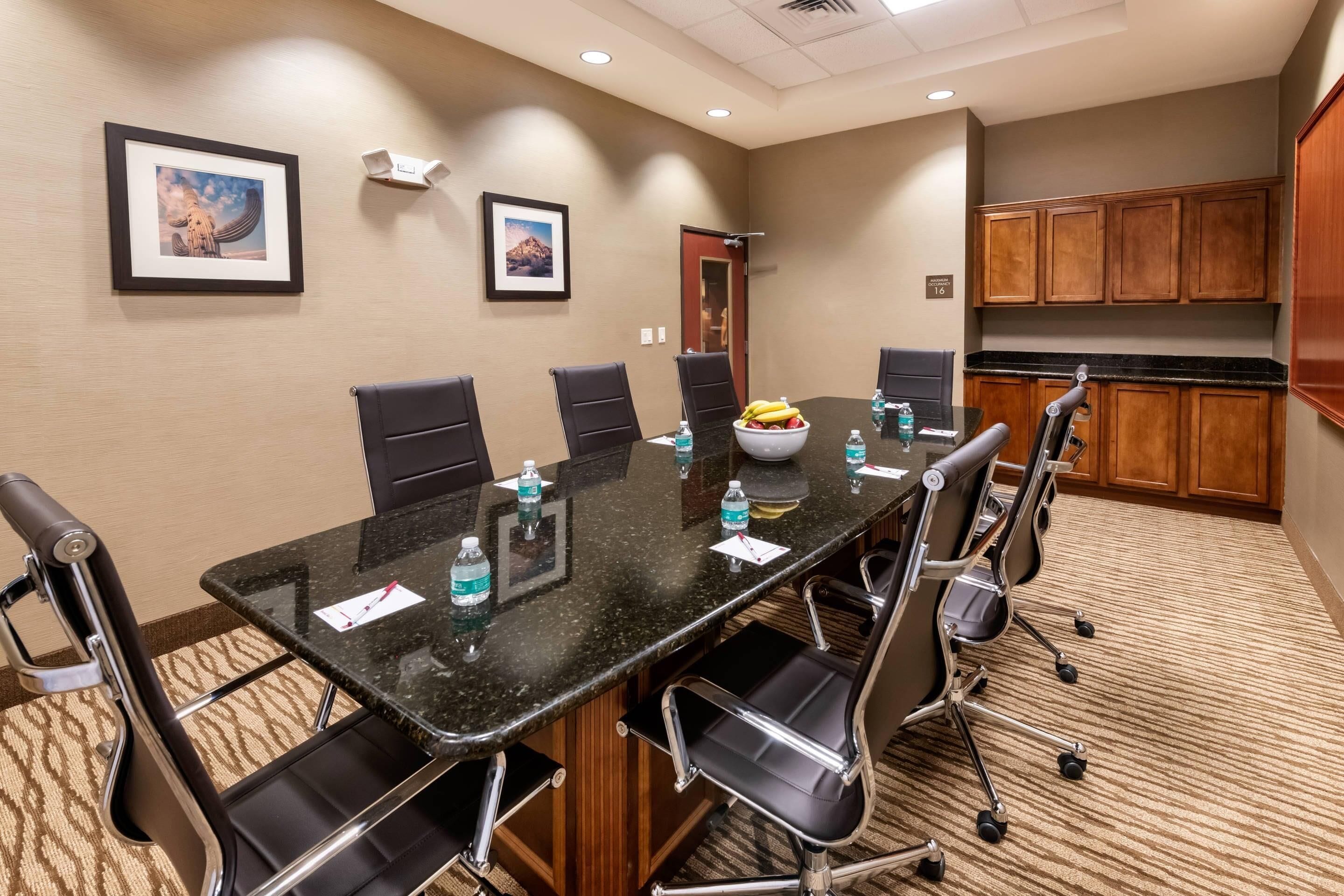 Uma sala de conferências com uma mesa comprida, seis cadeiras, quadros nas paredes e uma área de cozinha ao fundo.