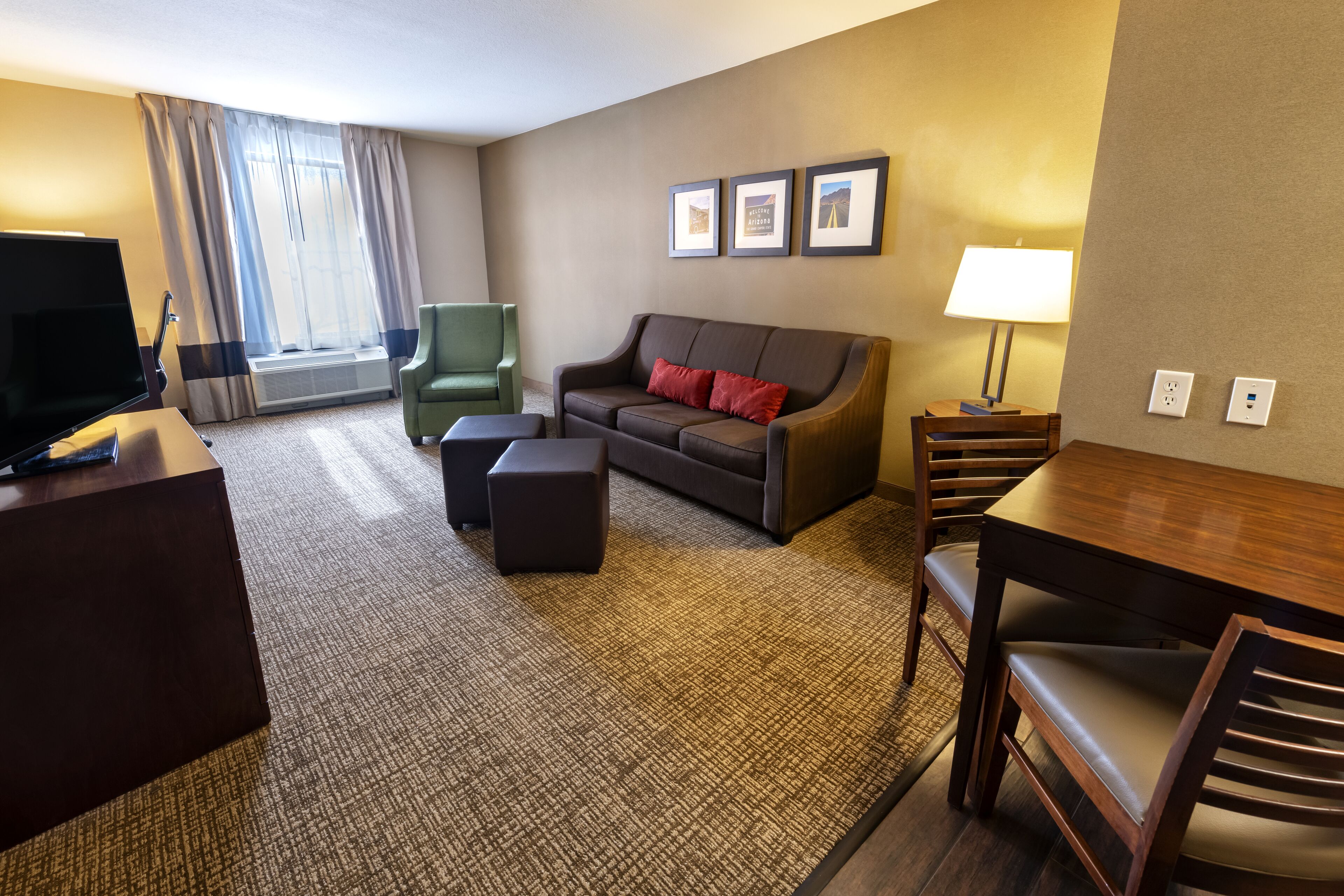 Quarto de hotel com sofá, televisão, mesa, cadeira, luminária e janela com cortinas.