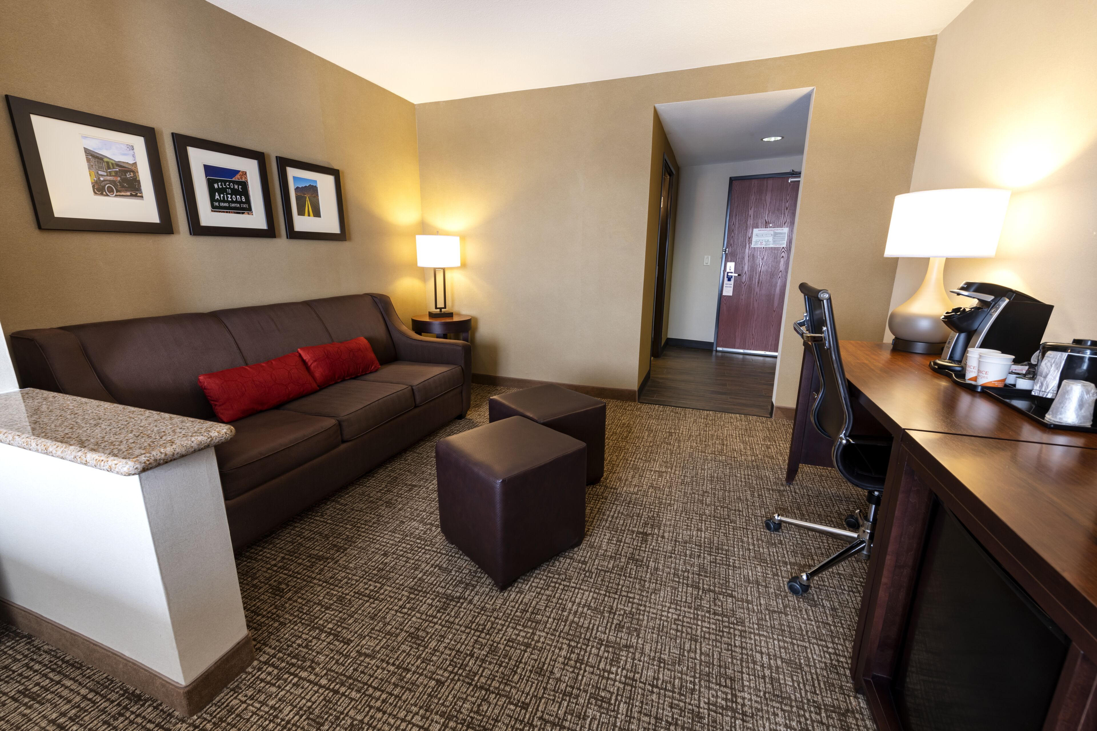Quarto de hotel com sofá de couro marrom, dois pufes escuros quadrados, uma pequena mesa lateral com abajur, escrivaninha com computador e cafeteira, e quadros nas paredes.