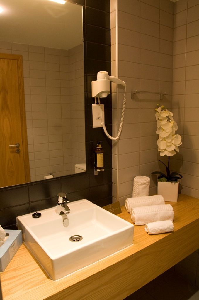 Casa de banho com lavatório branco, espelho, secador de cabelo, porta-toalhas e uma orquídea em vaso.