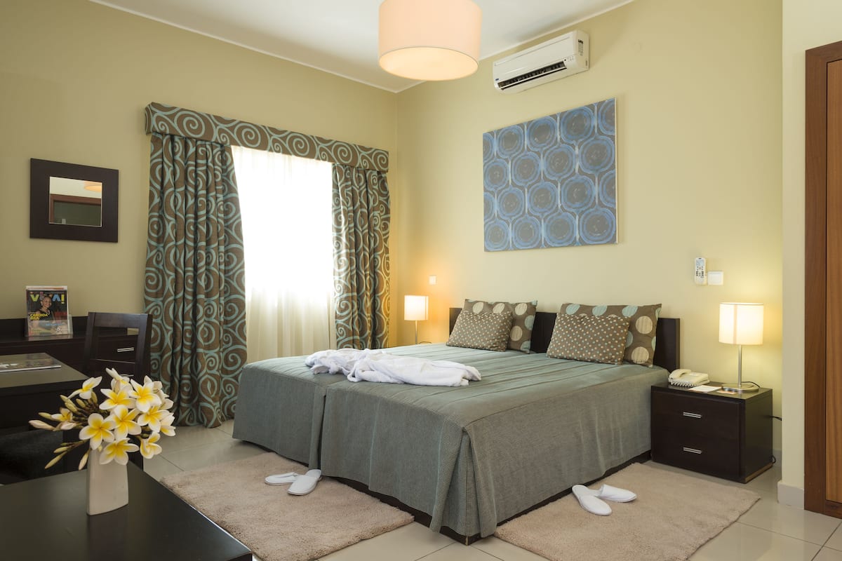 Quarto de hotel com cama de casal, duas mesas de cabeceira, escrivaninha, cadeira, vaso com flores e uma janela grande com cortinas.