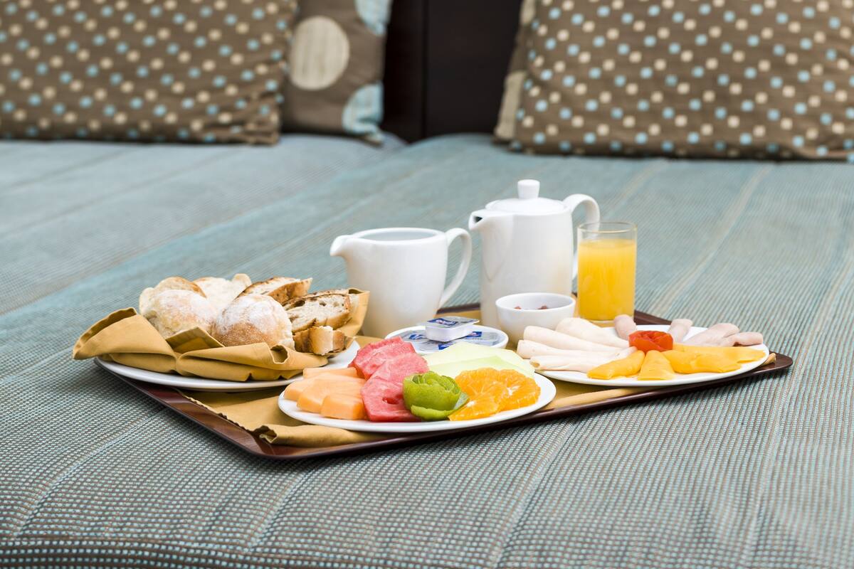 Uma bandeja de café da manhã com torradas, ovos, frutas e suco sobre uma mesa com toalha de mesa estampada em azul.
