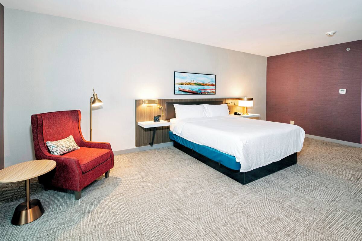 Quarto de hotel com cama, uma poltrona vermelha, uma mesa redonda e duas luminárias de cabeceira.