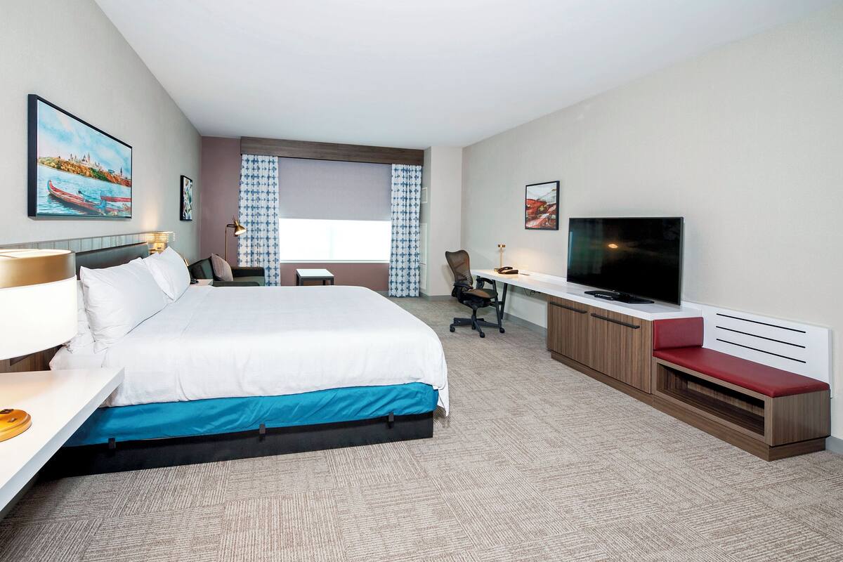 Quarto de hotel com cama, escrivaninha, televisão e janela com cortinas.