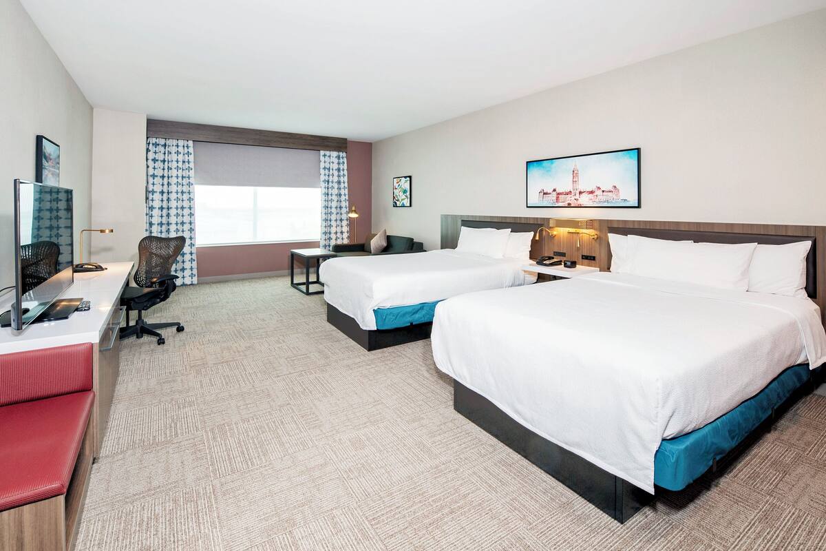 Quarto de hotel com duas camas, uma escrivaninha com computador, uma cadeira e um sofá.