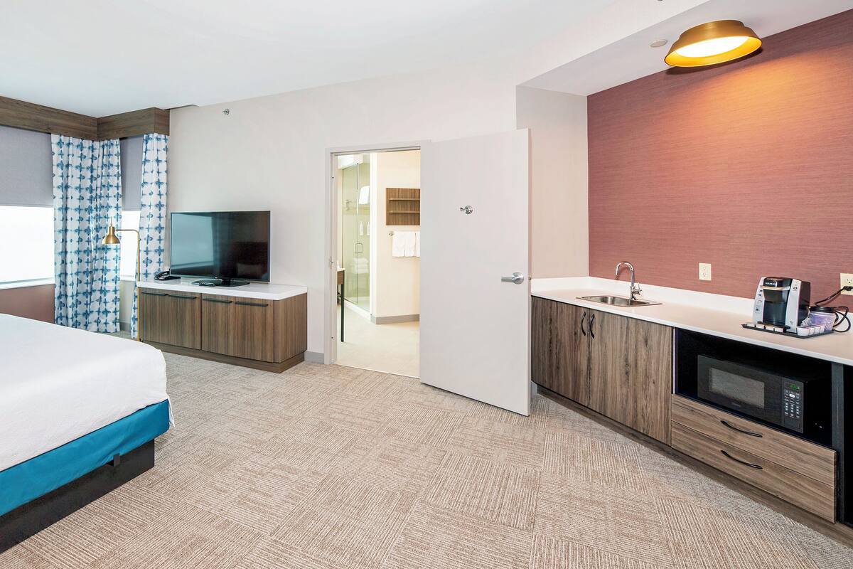 Quarto de hotel com cama, televisão, cozinha compacta e banheiro visível por uma porta aberta.