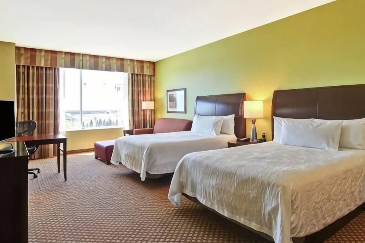 Quarto de hotel com duas camas, uma escrivaninha, uma cadeira, televisão e janela com cortinas.