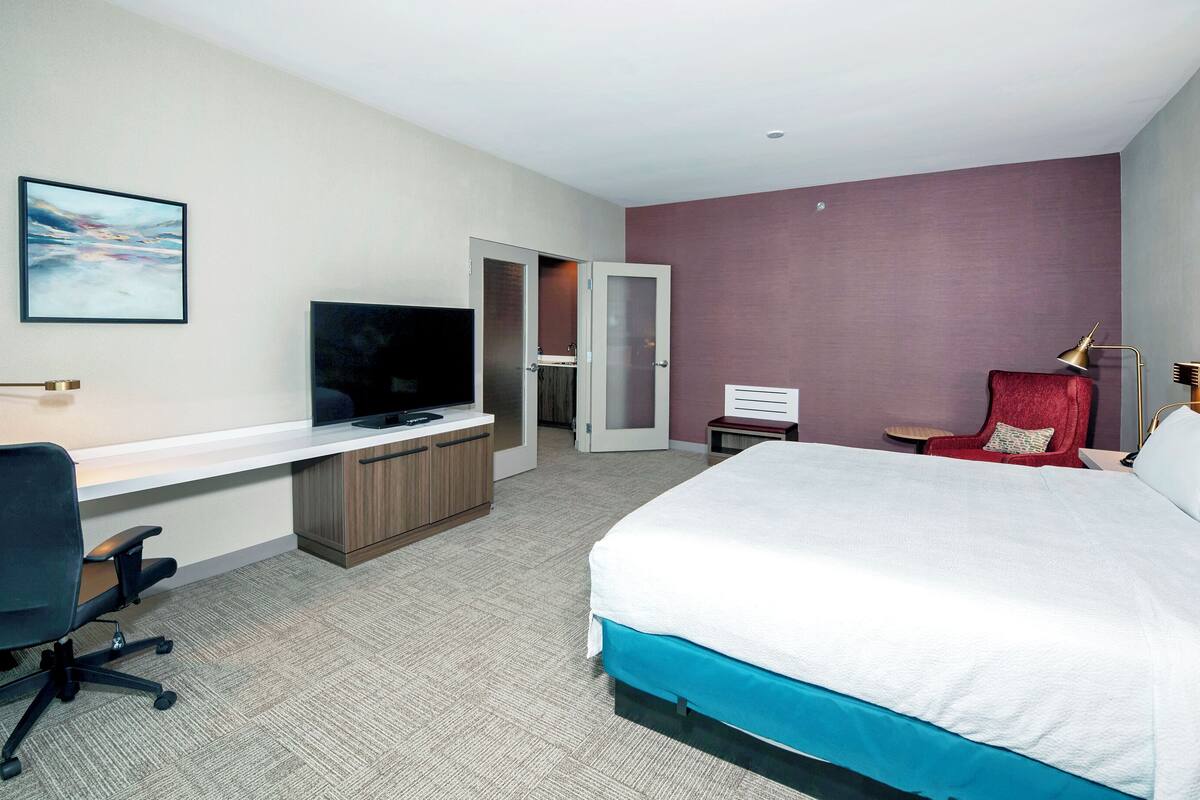 Quarto de hotel com cama, escrivaninha, cadeira, televisão e porta para um banheiro.